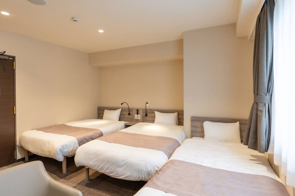 Quarto de hotel com três camas dispostas em linha, um guarda-roupa de madeira e uma escrivaninha com cadeira.