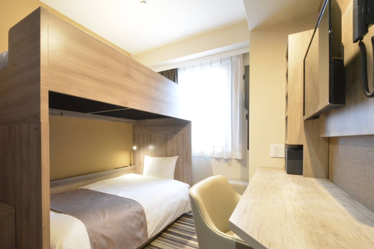 Quarto de hotel com uma cama beliche, uma cama de solteiro, uma mesa e uma cadeira.