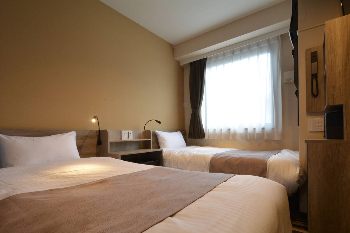 Quarto de hotel com duas camas, uma mesa, uma lâmpada e uma janela com cortinas.