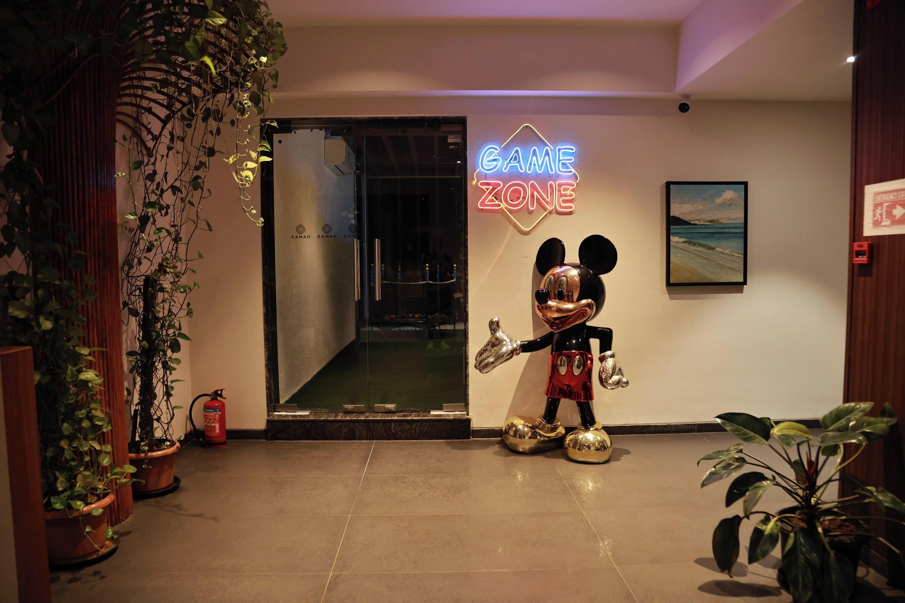 Entrada de hotel com um letreiro de neon que diz 'GAME ZONE', uma estátua do Mickey Mouse e um extintor de incêndio.