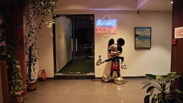 Entrada de hotel com um letreiro de neon que diz 'GAME ZONE', uma estátua do Mickey Mouse e um extintor de incêndio.