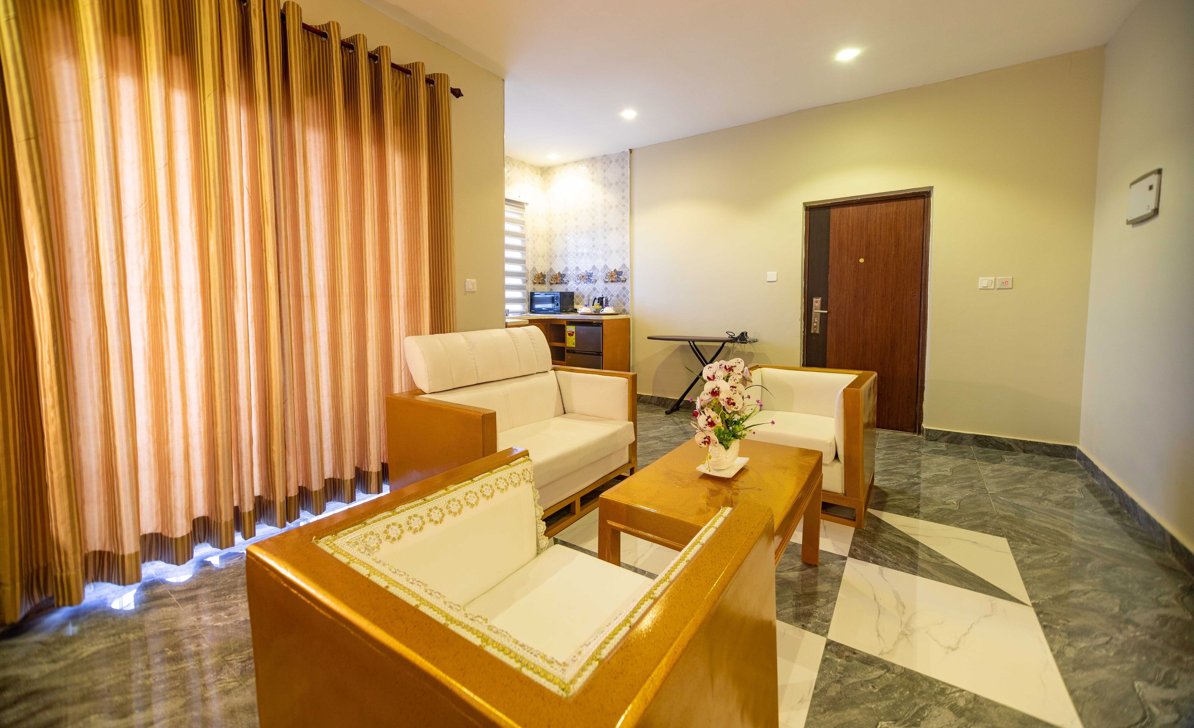 Uma sala de estar moderna com sofá, mesa de centro e mesa de jantar. Há uma área de cozinha com micro-ondas e pia. O espaço possui uma porta de madeira e uma janela com cortinas.