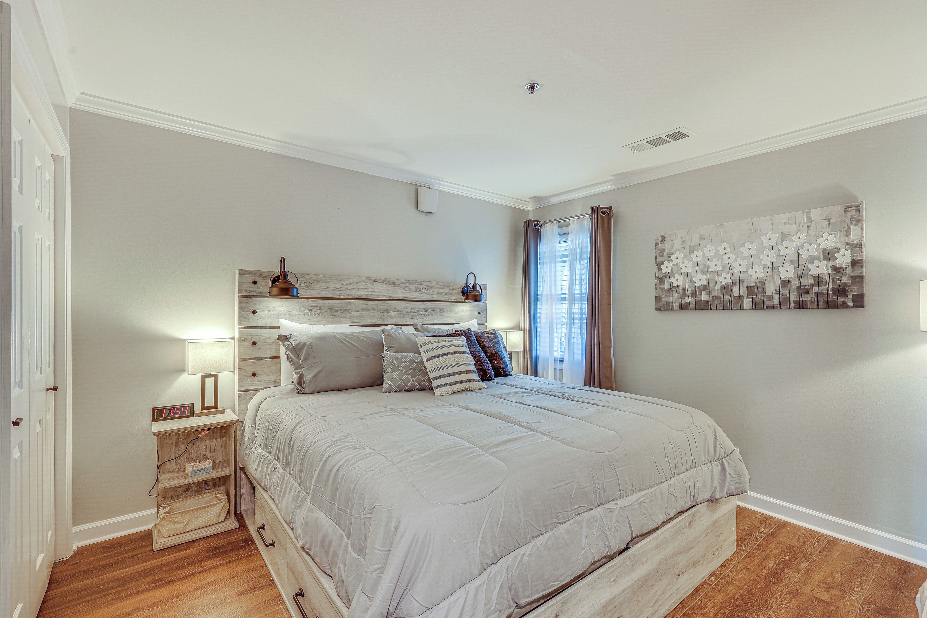 Quarto com cama grande, mesa de cabeceira com luminária, cabeceira de madeira e obra de arte na parede.