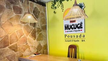 Balcão de recepção com parede de pedra ao fundo, luminária pendente e placa na parede com a inscrição 'Pousada Arrial d'Ajuada - BA'.