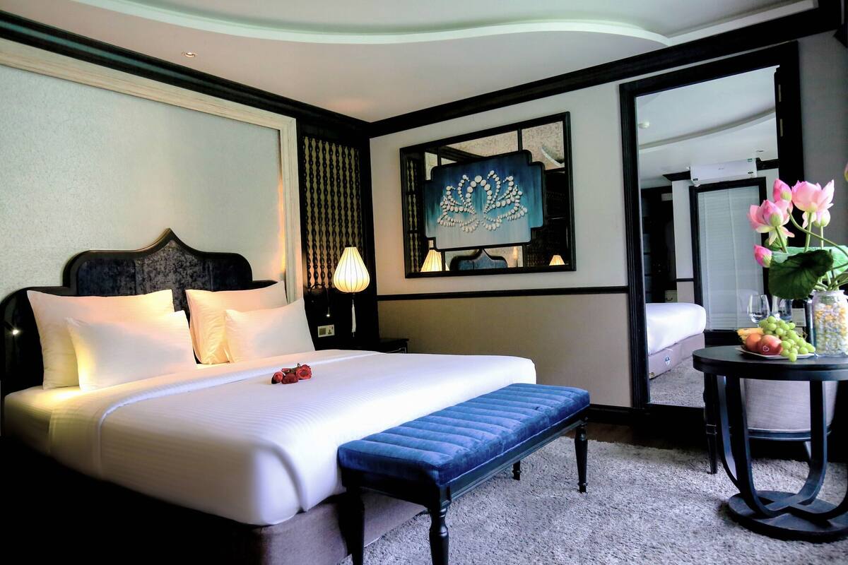 Cama bem arrumada com lençóis brancos e um banco azul, uma pequena mesa com um vaso de flores e um quadro pendurado na parede.