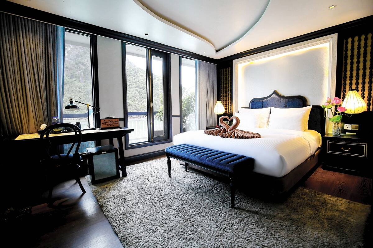 Quarto de hotel com uma cama grande, uma escrivaninha, uma cadeira e vista para o exterior através de amplas janelas.