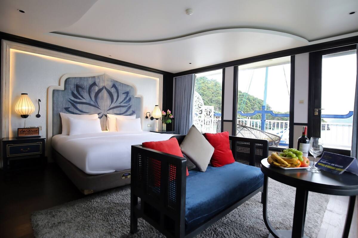 Quarto de hotel com cama, sofá, mesinha com fruta e uma taça de vinho, e vista para o mar.
