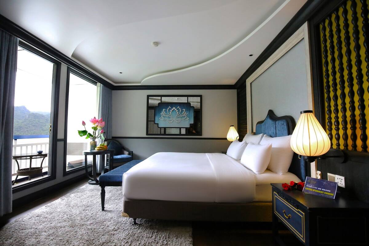 Quarto de hotel com uma cama grande, uma mesa de cabeceira com uma luminária, uma mesinha com um vaso de flores e vista para as montanhas pela janela.