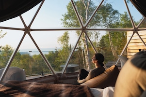 Quarto com uma janela grande, vista para árvores e uma pessoa sentada no sofá.