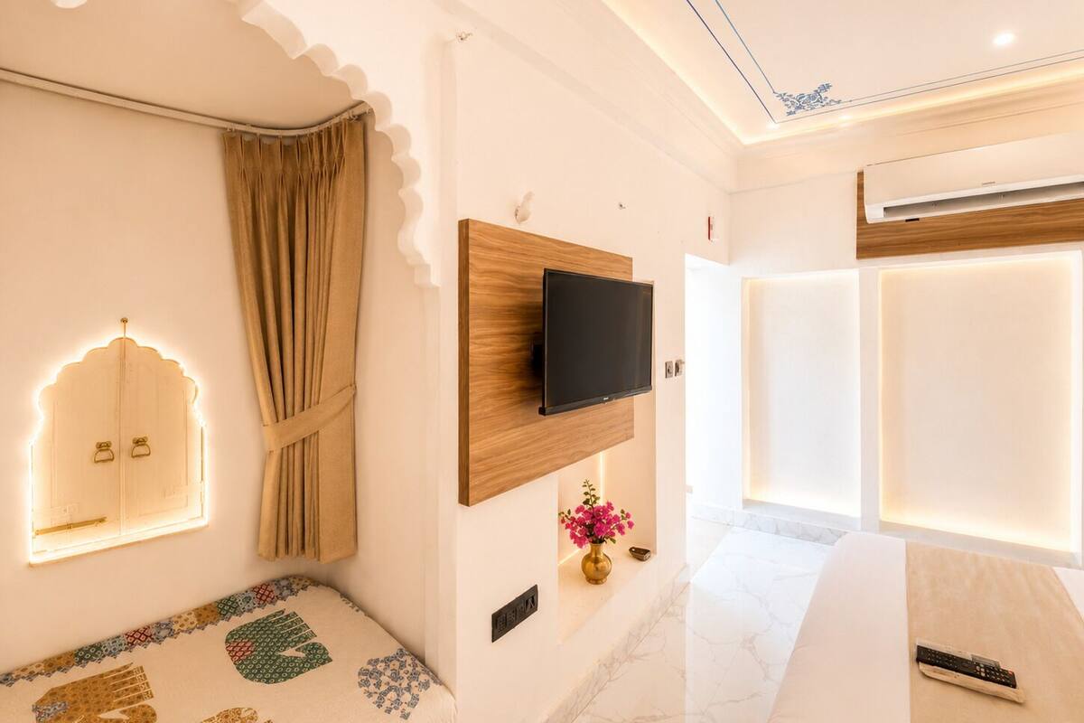 Quarto de hotel moderno com televisão de tela plana na parede, cama com cabeceira estampada, mesa pequena com um vaso de flores e janela com cortinas.