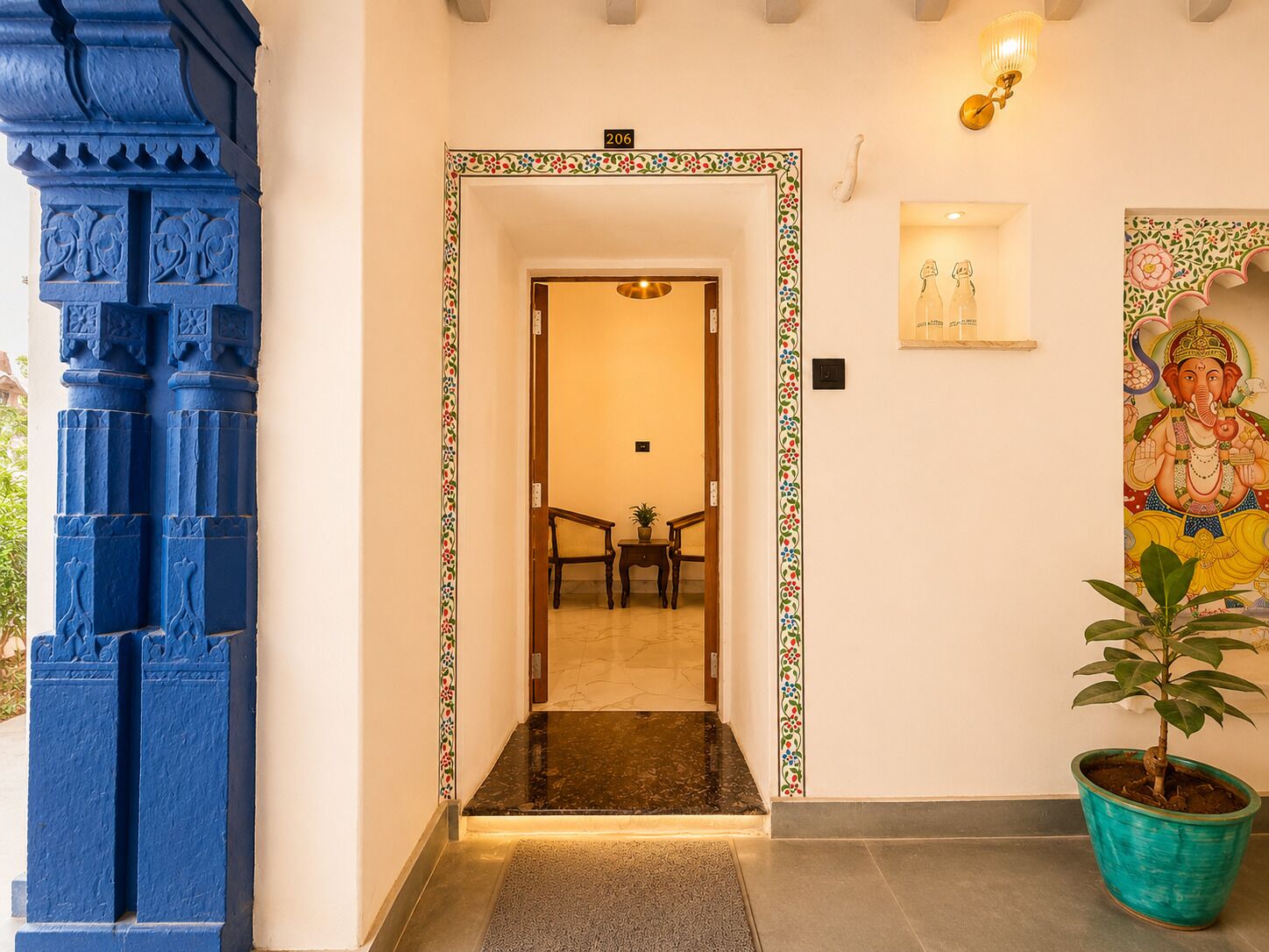 Um corredor com uma porta identificada com o número '206', uma planta em vaso e uma parede com uma pintura decorativa.
