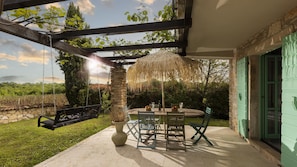 Um pátio externo com um balanço, uma mesa redonda com cadeiras, um guarda-sol de palha e uma parede de pedra ao fundo.