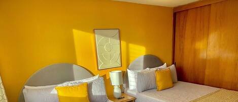Quarto de hotel com duas camas, um guarda-roupa de madeira, uma mesa de cabeceira com abajur e uma obra de arte na parede.