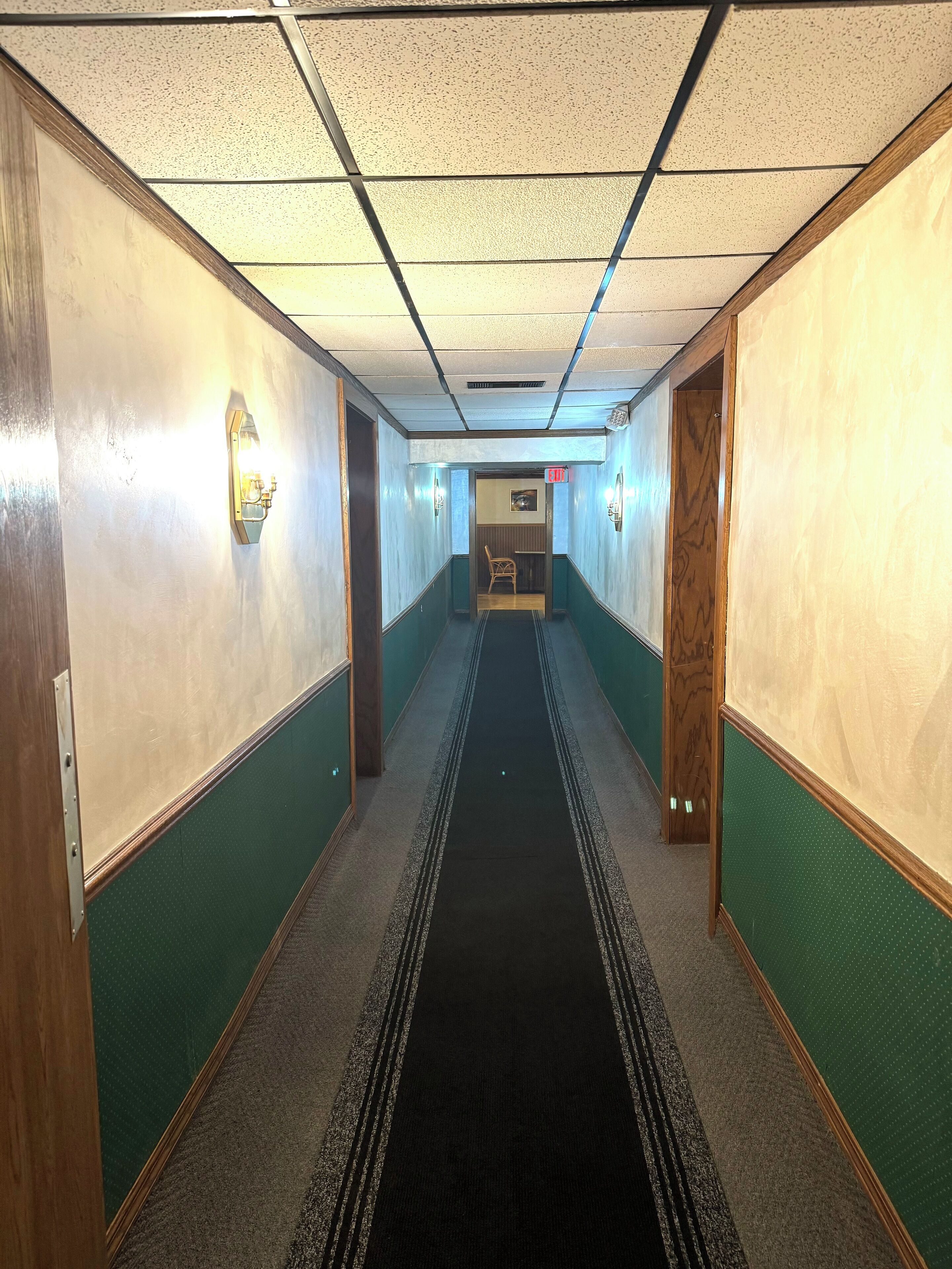 Um corredor de hotel com paredes verdes e bege, carpete preto e uma porta de madeira no final.