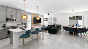 Uma cozinha moderna com uma ilha central, banquetas de bar, sala de jantar com mesa redonda e cadeiras, e área de estar com sofá e televisão.