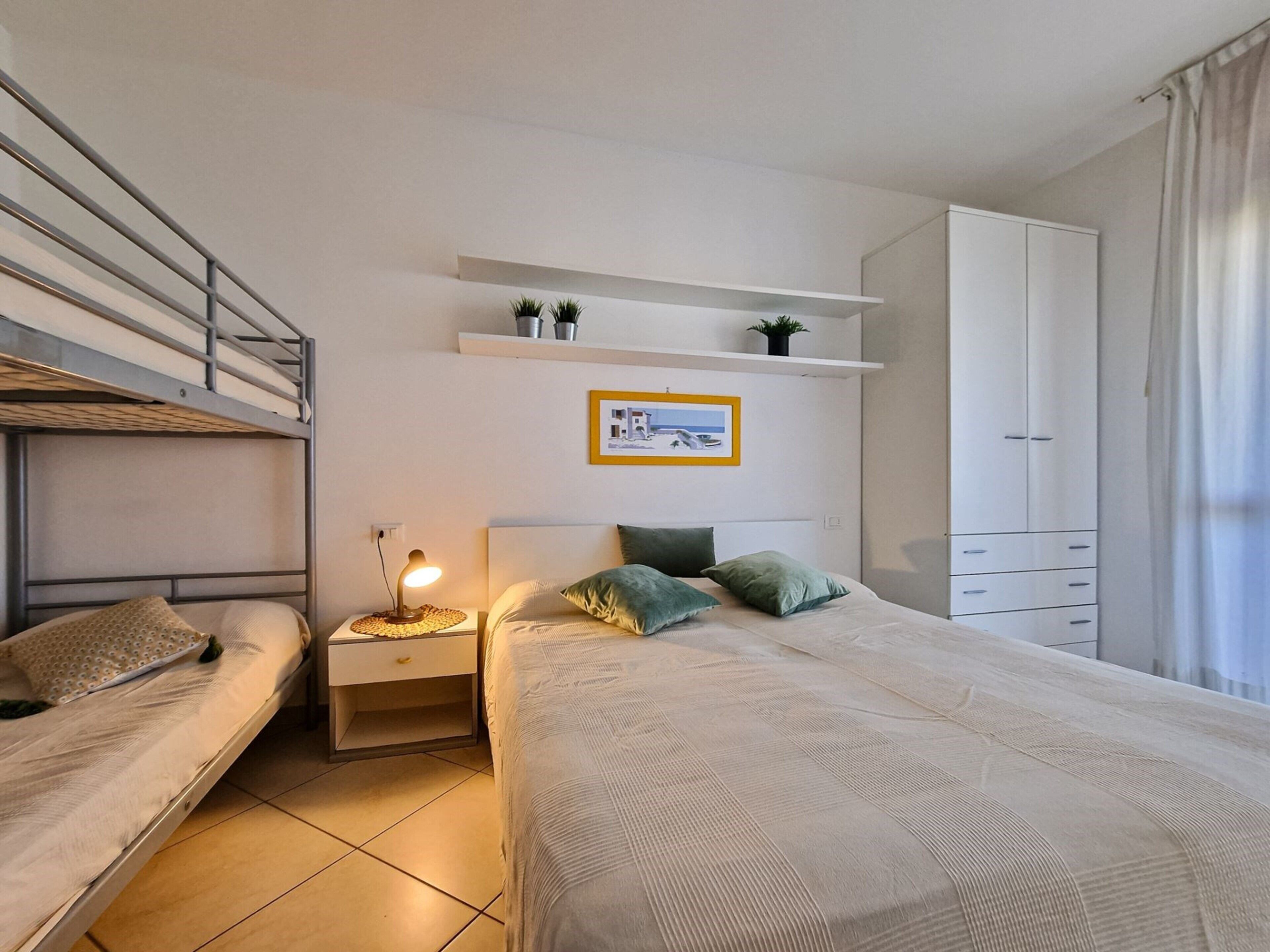 Quarto com beliches, mesa de cabeceira com abajur, cama com almofadas, prateleira com objetos decorativos, guarda-roupa e um quadro na parede.