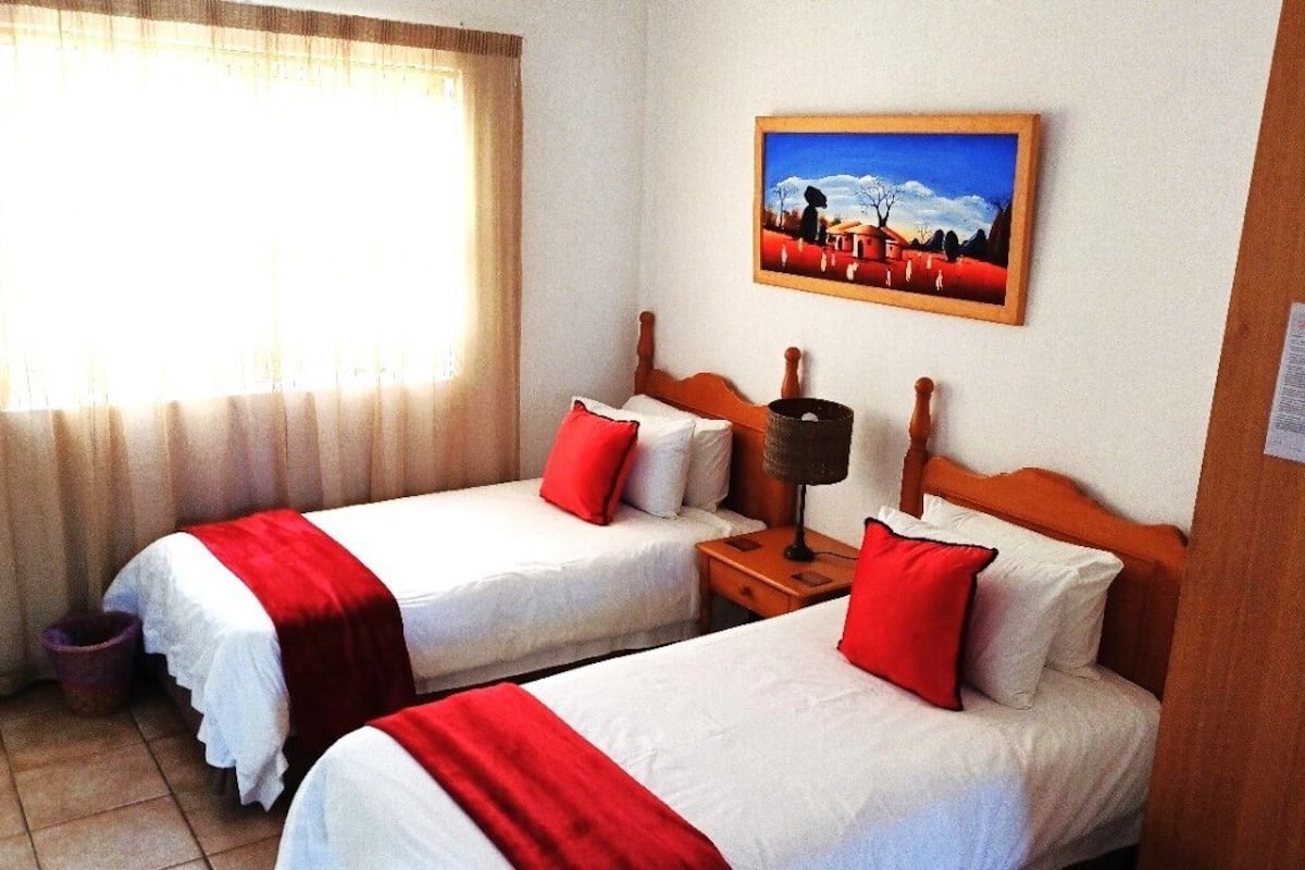 Quarto de hotel com duas camas, cada uma com colcha vermelha e branca, uma mesinha de cabeceira com abajur e um quadro na parede.