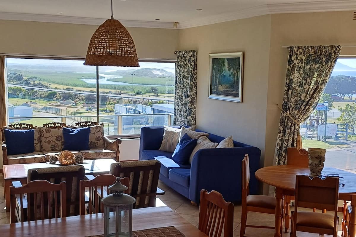 Uma sala de estar com sofá, mesa de jantar e cadeiras, uma janela grande com vista para uma paisagem, e duas luminárias pendentes.