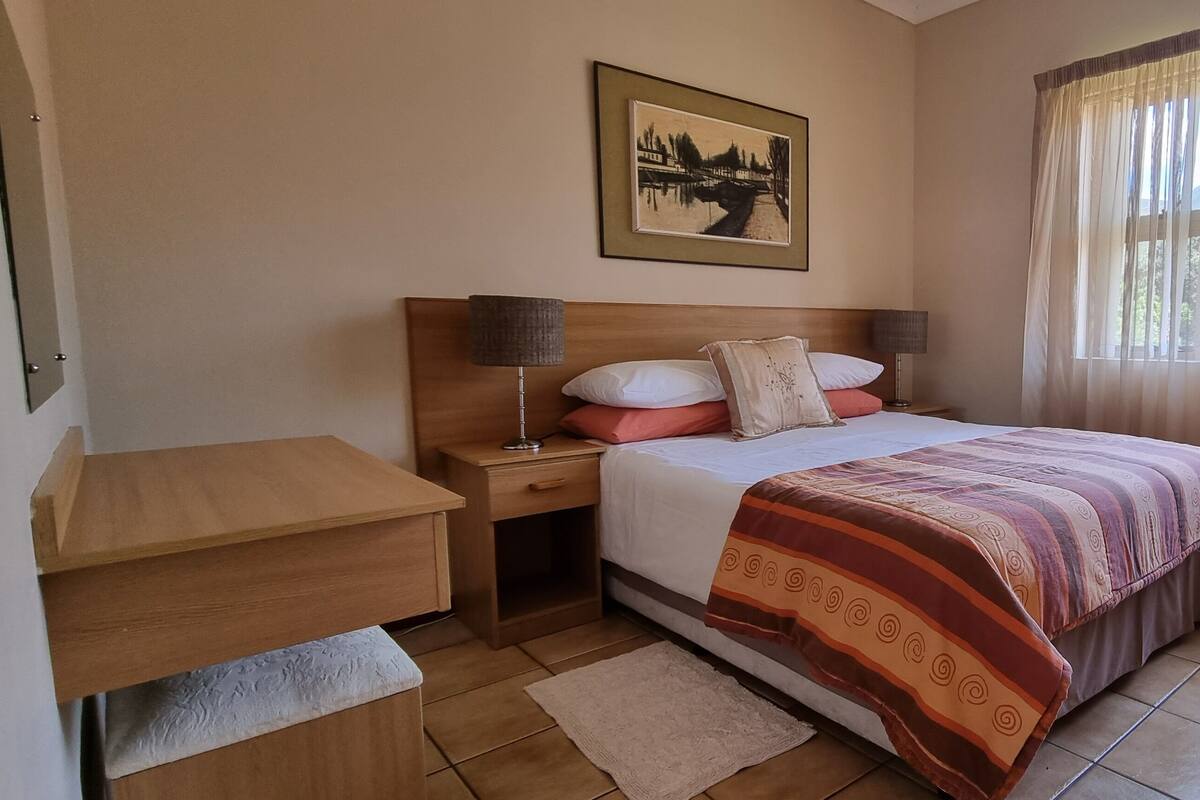 Quarto com cama, mesas de cabeceira, luminária, quadro pendurado na parede e janela com cortinas.