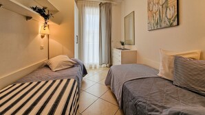 Quarto de hotel com duas camas, uma mesa de cabeceira com abajur, espelho, um quadro na parede e janela com cortinas.