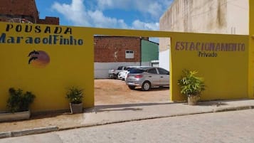 Um portão amarelo com placas indicando "Pousada Maracolinho" e "Estacionamento (Privado)", dando acesso a uma área de estacionamento com carros.