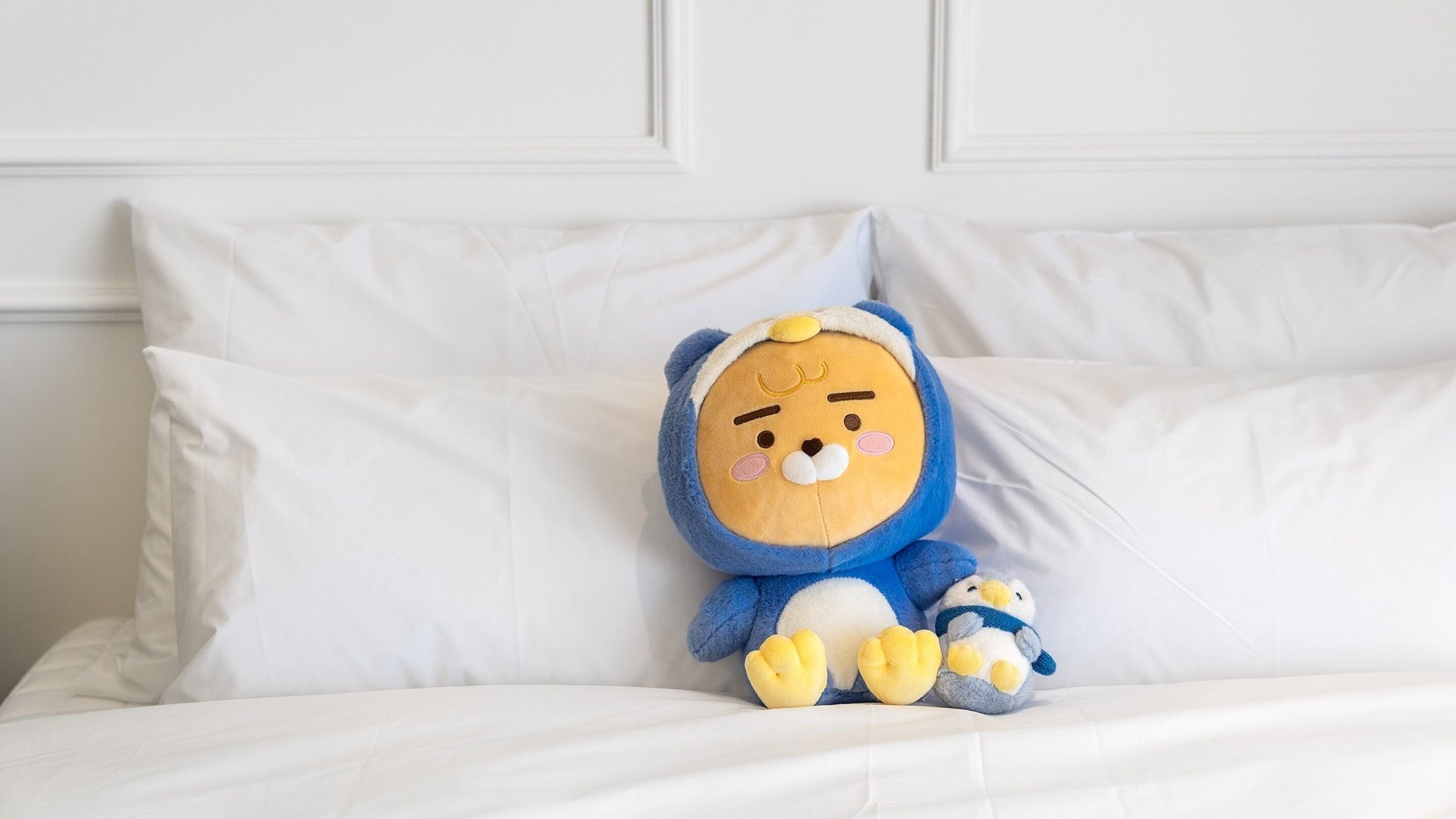 Uma cama com roupa de cama e almofadas brancas, um brinquedo de pelúcia com roupa azul e amarela, e um brinquedo menor ao lado.