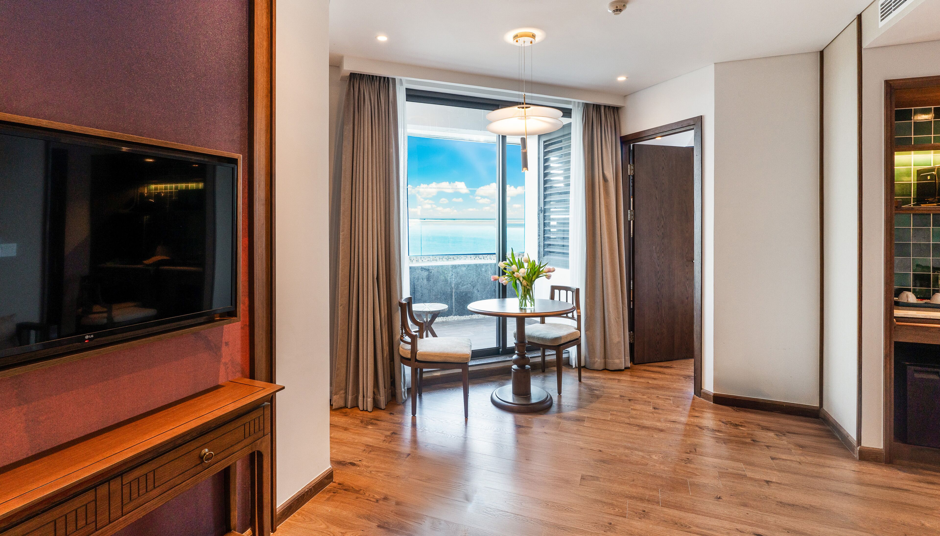 Quarto de hotel com televisão, área de refeições com mesa e cadeiras, vista para o oceano e piso de madeira.