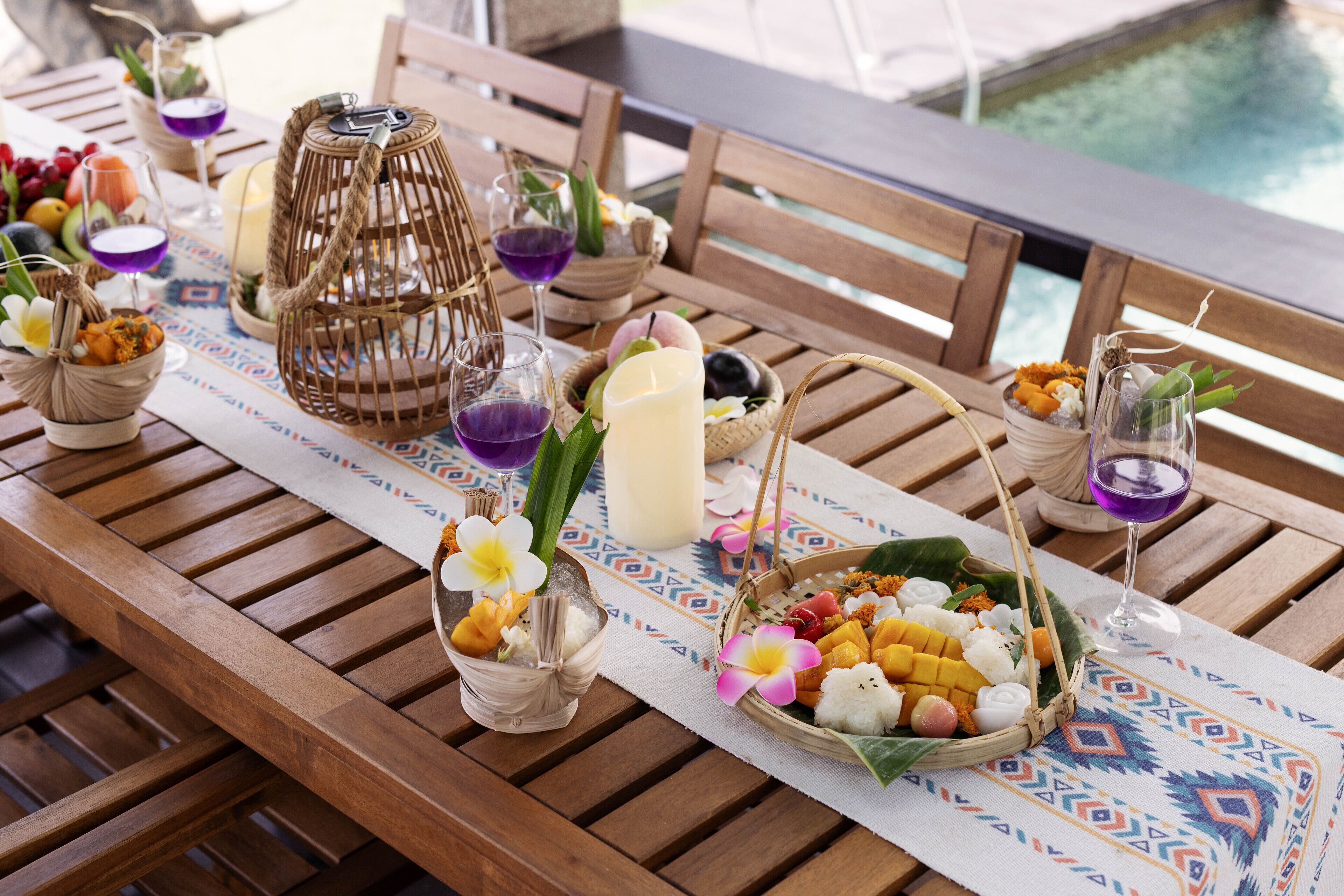 Uma mesa de madeira com uma cesta de palha contendo frutas e uma vela, rodeada por objetos decorativos e uma toalha de mesa estampada.
