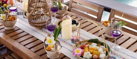 Uma mesa de madeira com uma cesta de palha contendo frutas e uma vela, rodeada por objetos decorativos e uma toalha de mesa estampada.