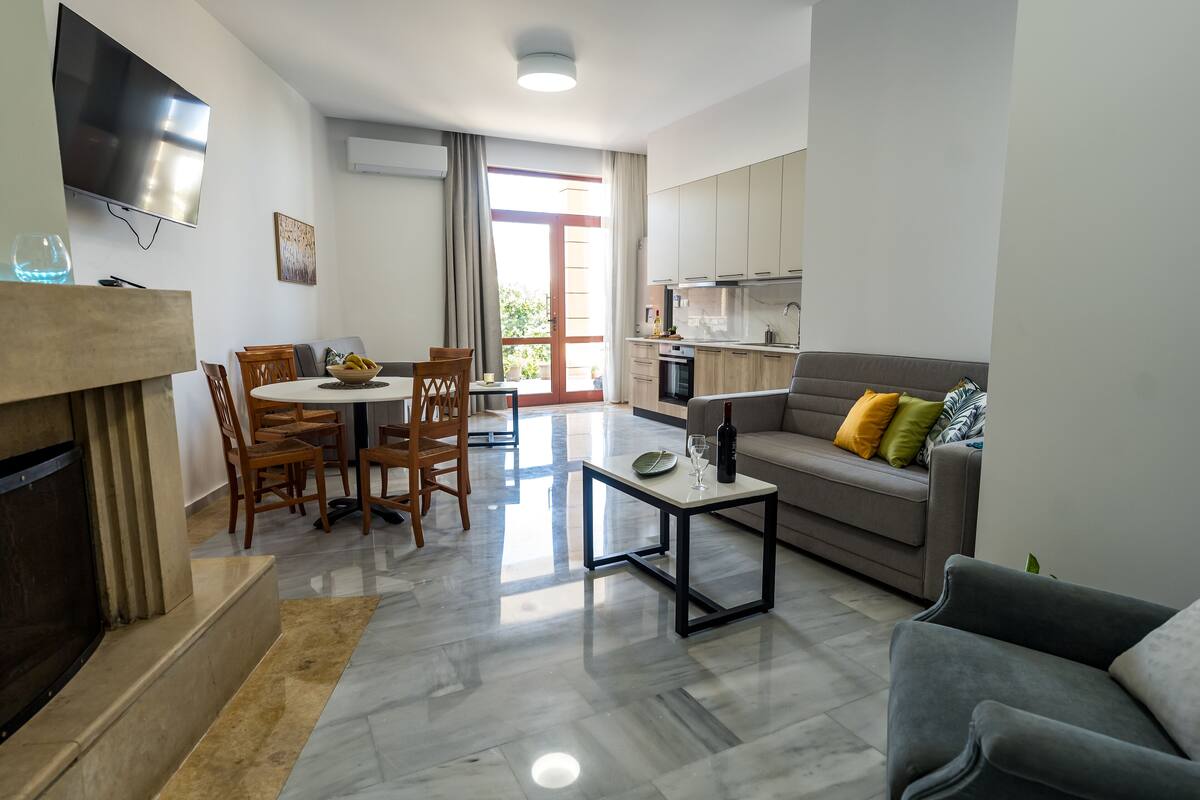 Uma sala de estar moderna com lareira, um espaço de jantar com mesa e cadeiras, um sofá e uma cozinha com armários e uma televisão.