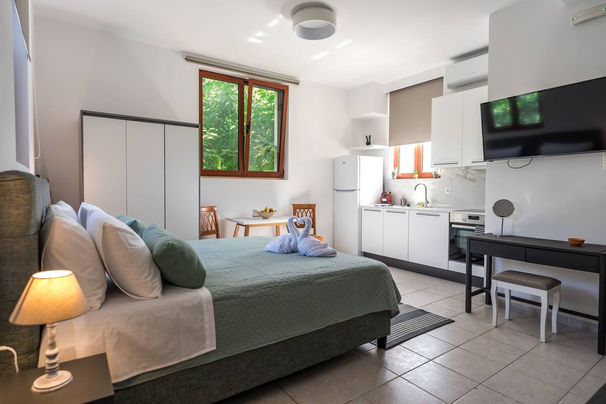 Quarto com cama, mesa de cabeceira, luminária e vista para uma cozinha com mesa e cadeiras.