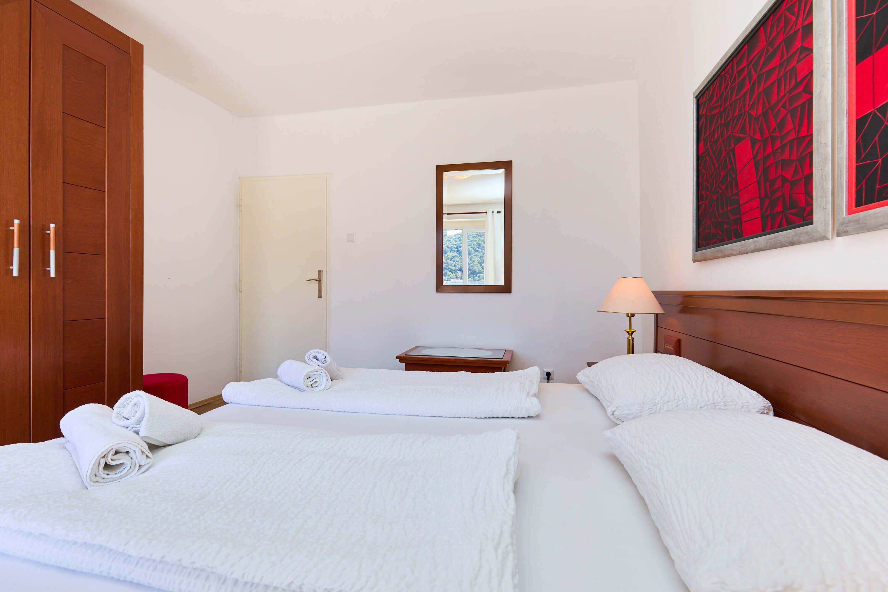 Quarto de hotel com duas camas, guarda-roupa de madeira, espelho, mesa de cabeceira com abajur e uma obra de arte emoldurada na parede.