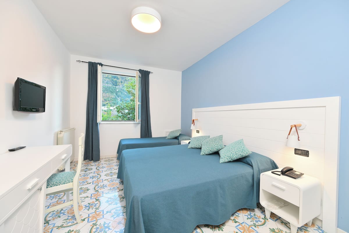 Quarto com duas camas, uma escrivaninha, uma cadeira, uma televisão e uma janela com cortinas.