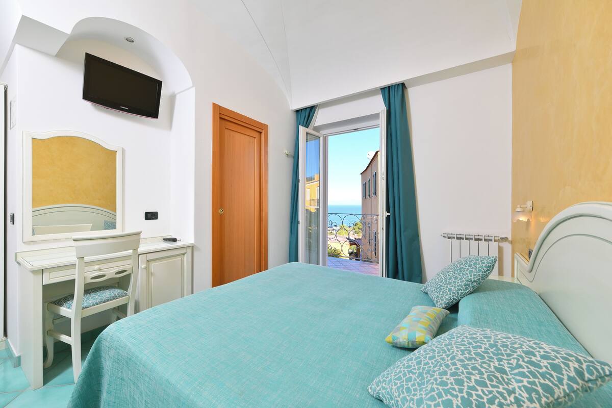 Quarto com cama, escrivaninha, cadeira, TV fixada na parede e varanda com vista para o mar.