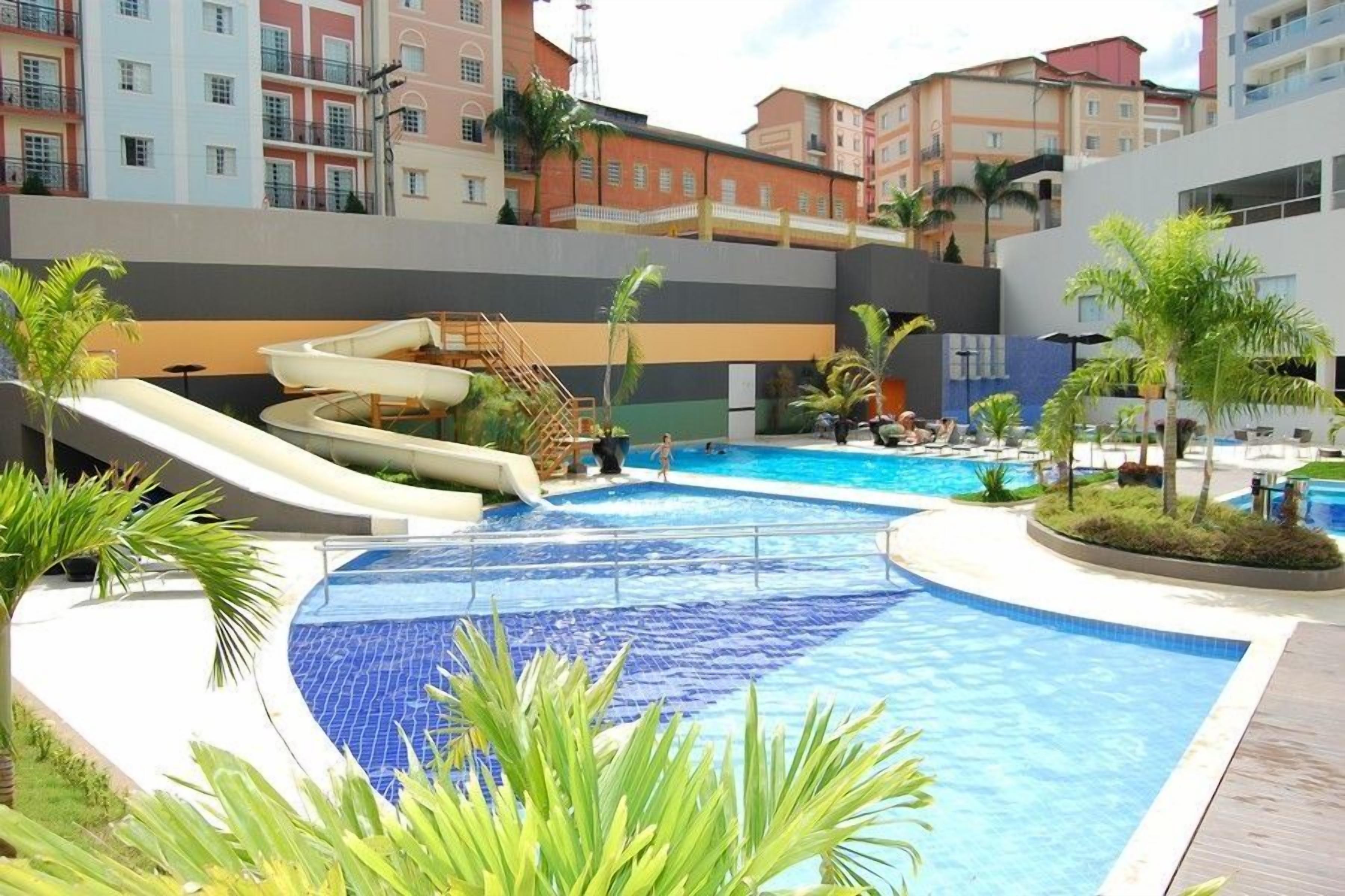 Piscina com escorregador, cercada por palmeiras e um edifício alto ao fundo.
