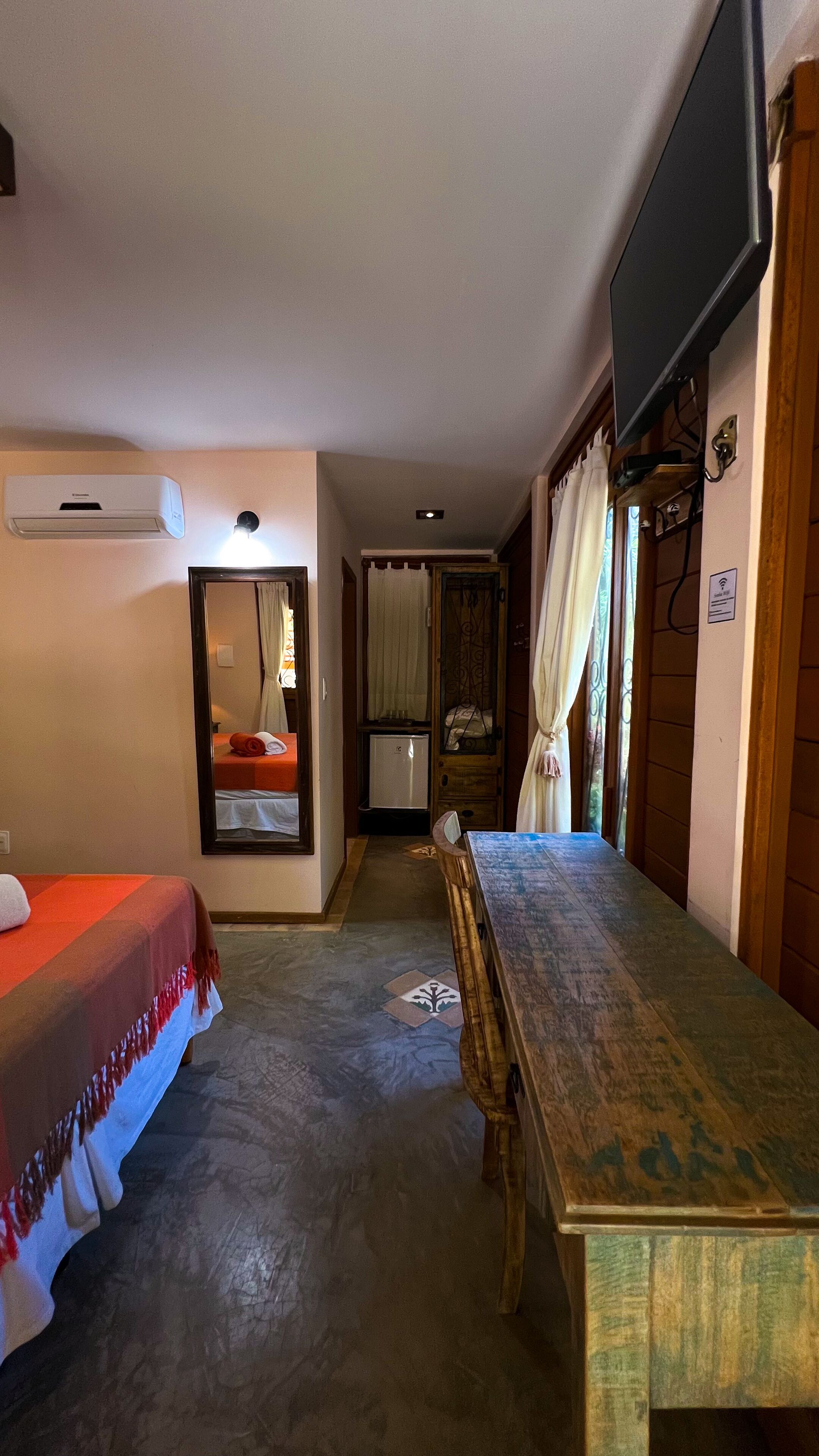 Quarto com cama, uma mesa pequena, espelho, televisão e frigorífico.