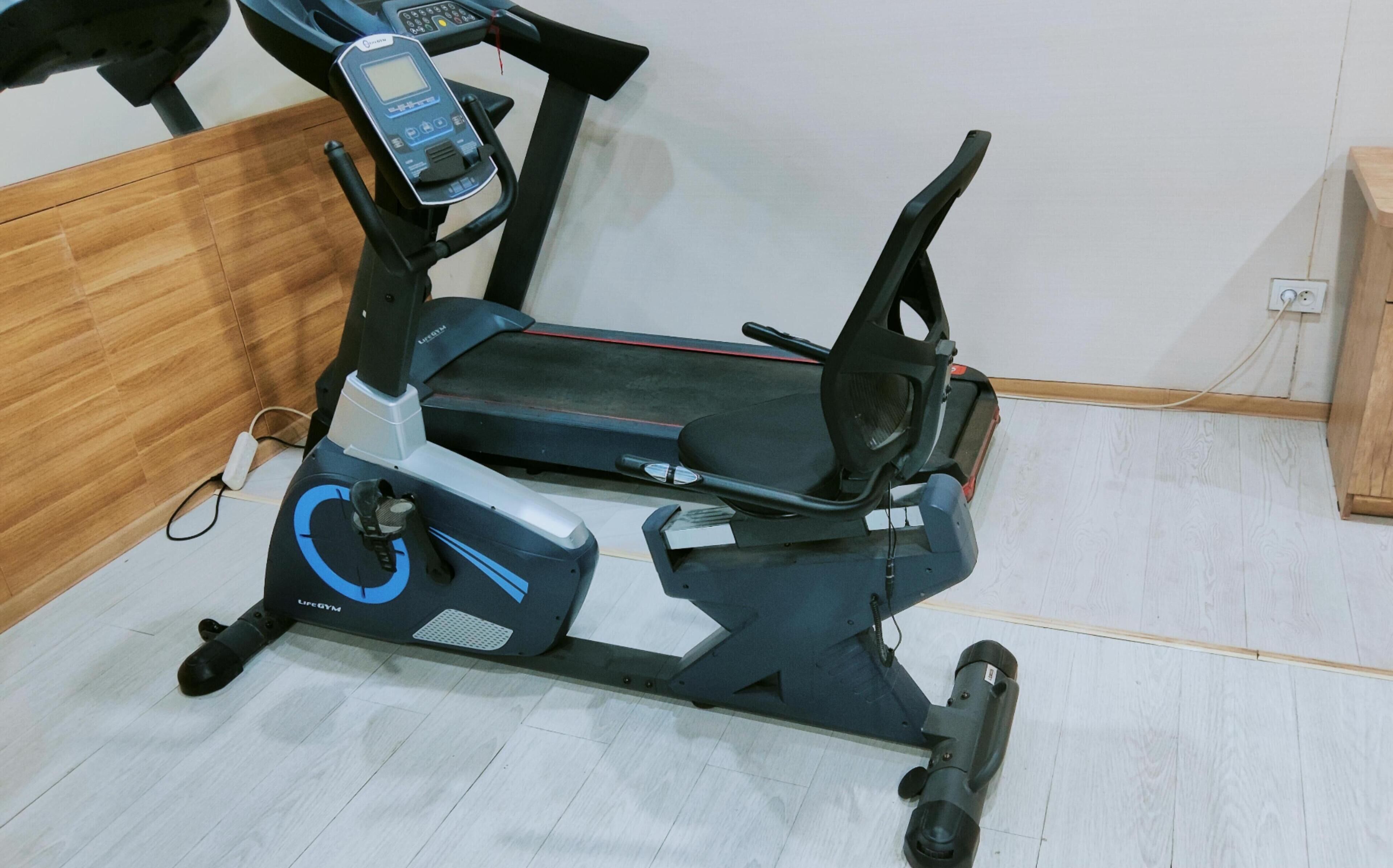 Uma bicicleta ergométrica com visor digital, painel de controle e cabo de alimentação conectado a uma tomada.