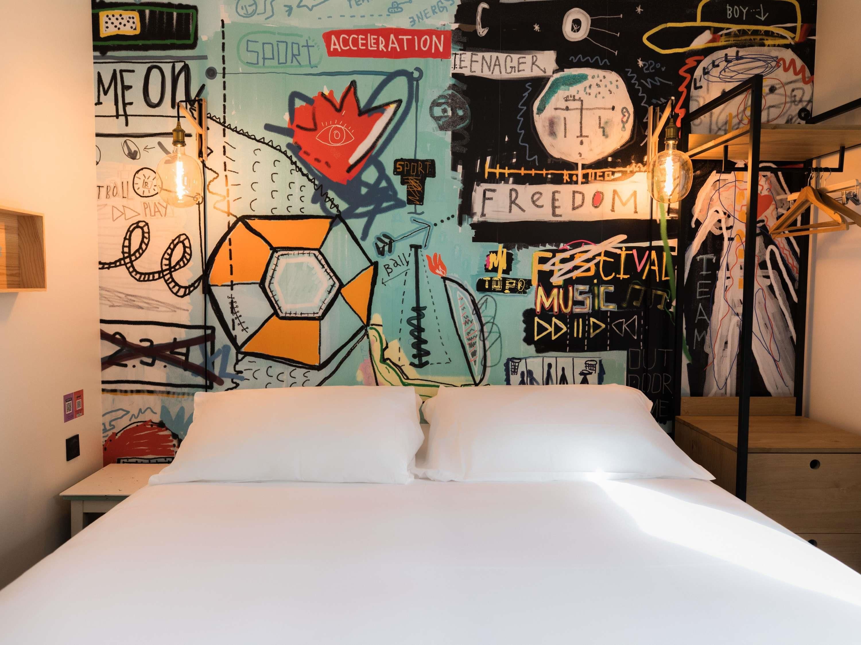 Quarto com cama, um mural na parede com várias tags e rabiscos, e uma prateleira de madeira com objetos pendurados.