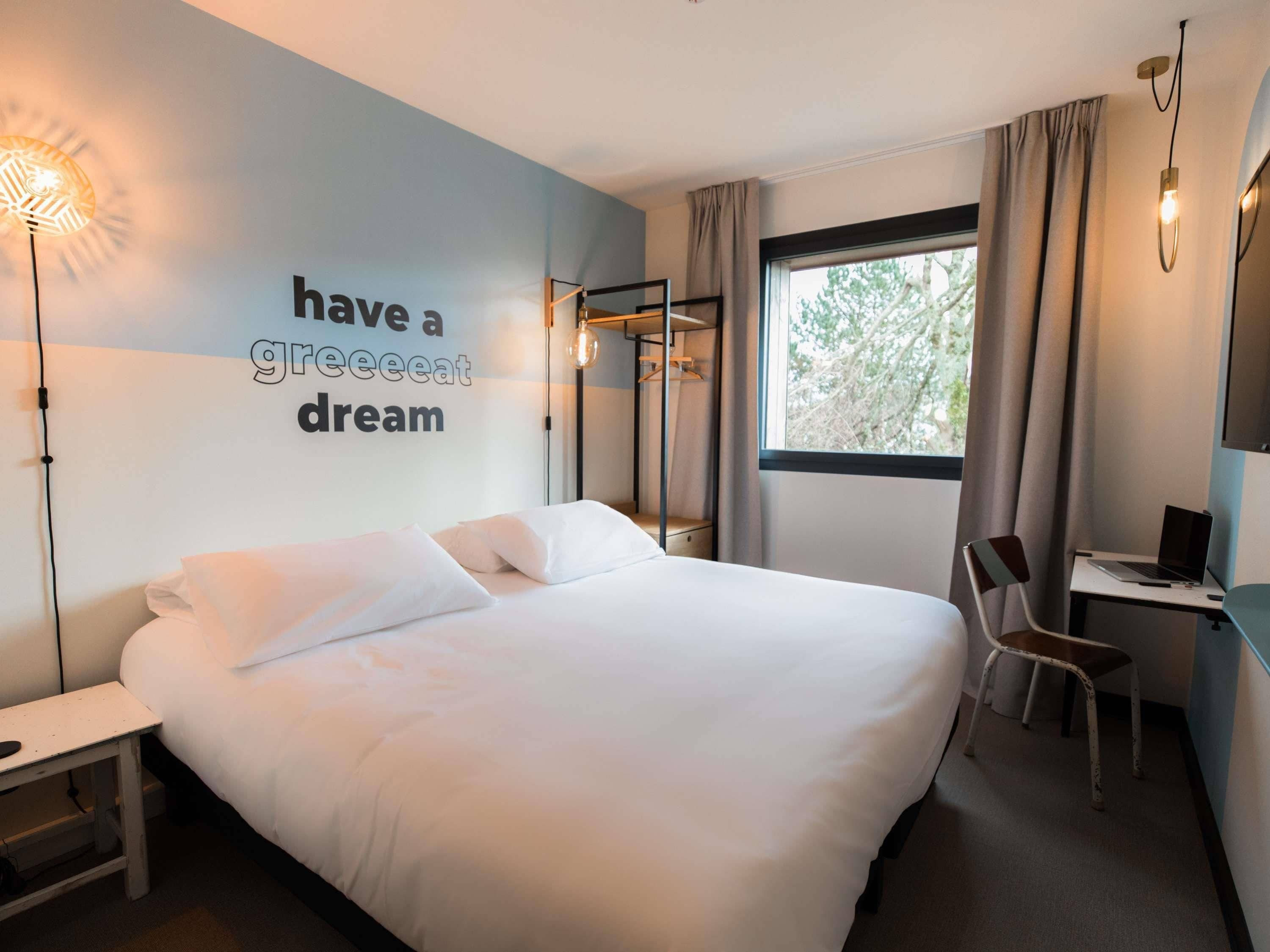 Quarto de hotel moderno com cama, escrivaninha, cadeira e uma frase motivacional na parede.