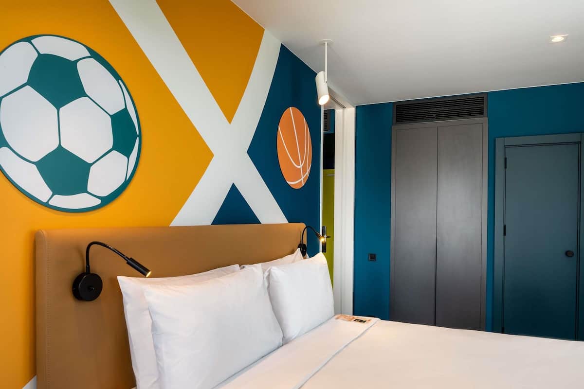 Quarto de hotel com uma cama, cabeceira, parede com um design geométrico grande, porta e luminária no teto.