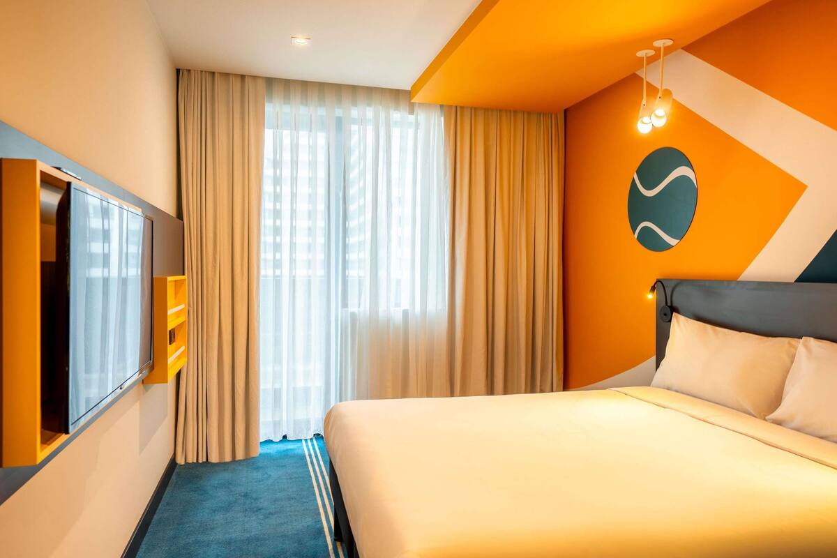 Quarto de hotel com cama, televisão, janela grande com cortinas e parede com design geométrico.