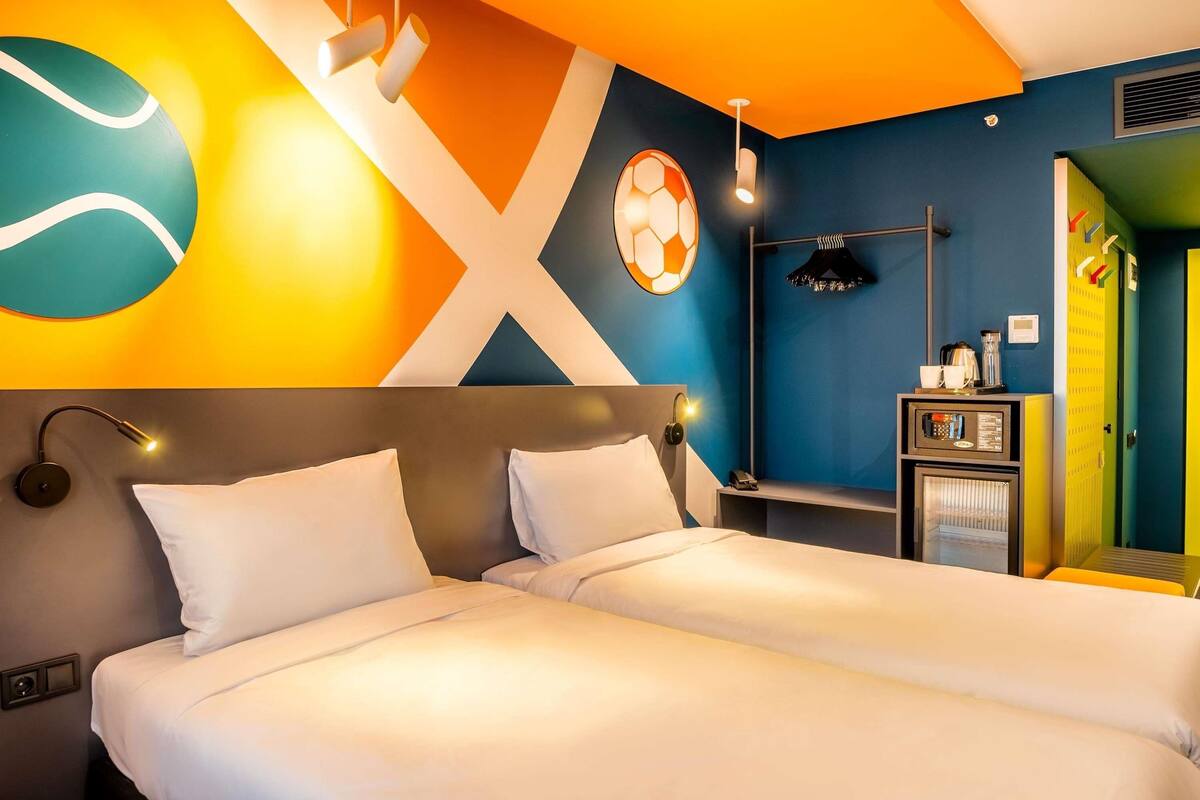 Quarto de hotel com cama, mesas de cabeceira, uma obra de arte na parede, luminária pendente e um porta-roupas.
