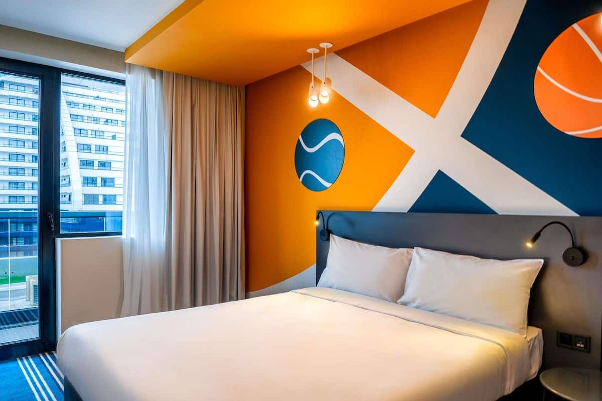 Quarto de hotel com uma cama grande, vista da cidade pela janela e um design de parede colorido.