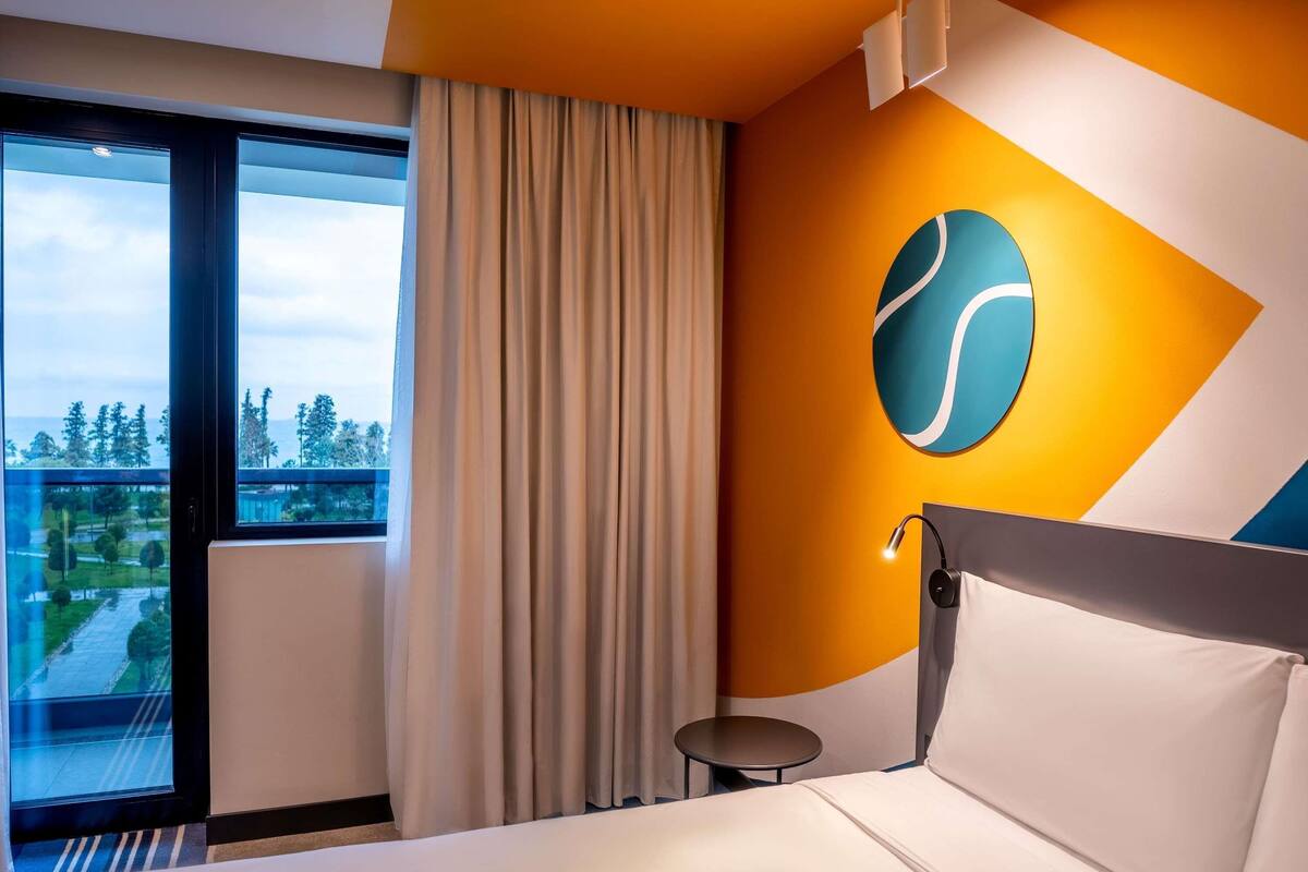 Quarto de hotel com uma cama, uma mesinha, cortina, janela com vista e parede com um desenho geométrico.