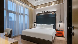 Quarto de hotel moderno com uma cama grande, uma mesa e uma cadeira. Há uma mesinha de cabeceira com uma luminária e um relógio. O quarto tem vista para o horizonte urbano pela janela.