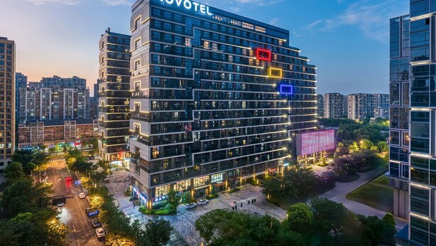 Um hotel moderno com um sinal grande 'NOVOTEL', rodeado por outros edifícios e uma área de estacionamento bem iluminada.