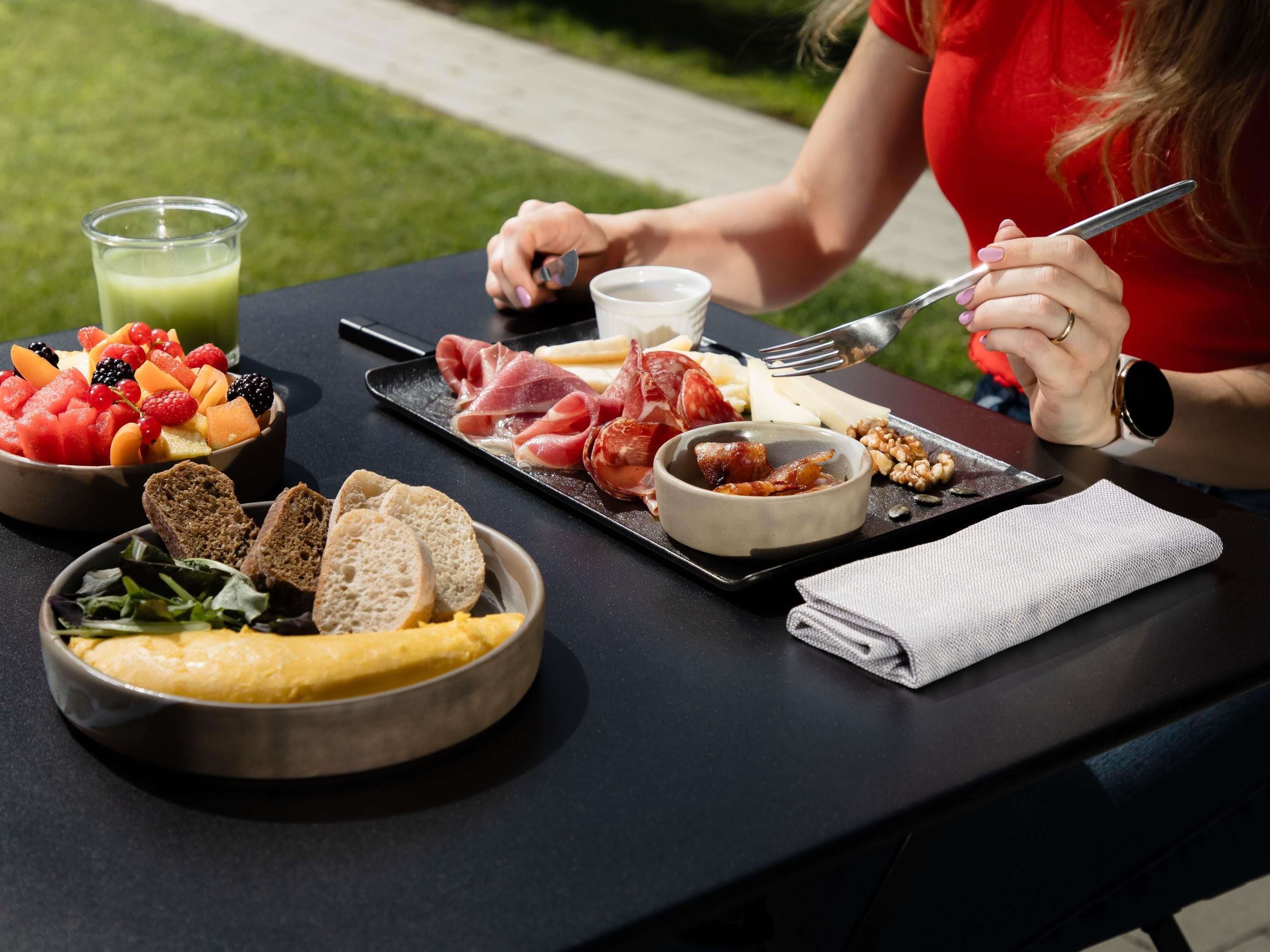 Uma refeição com diversos pratos, incluindo frutas, carnes e pão, servida em uma mesa ao ar livre.