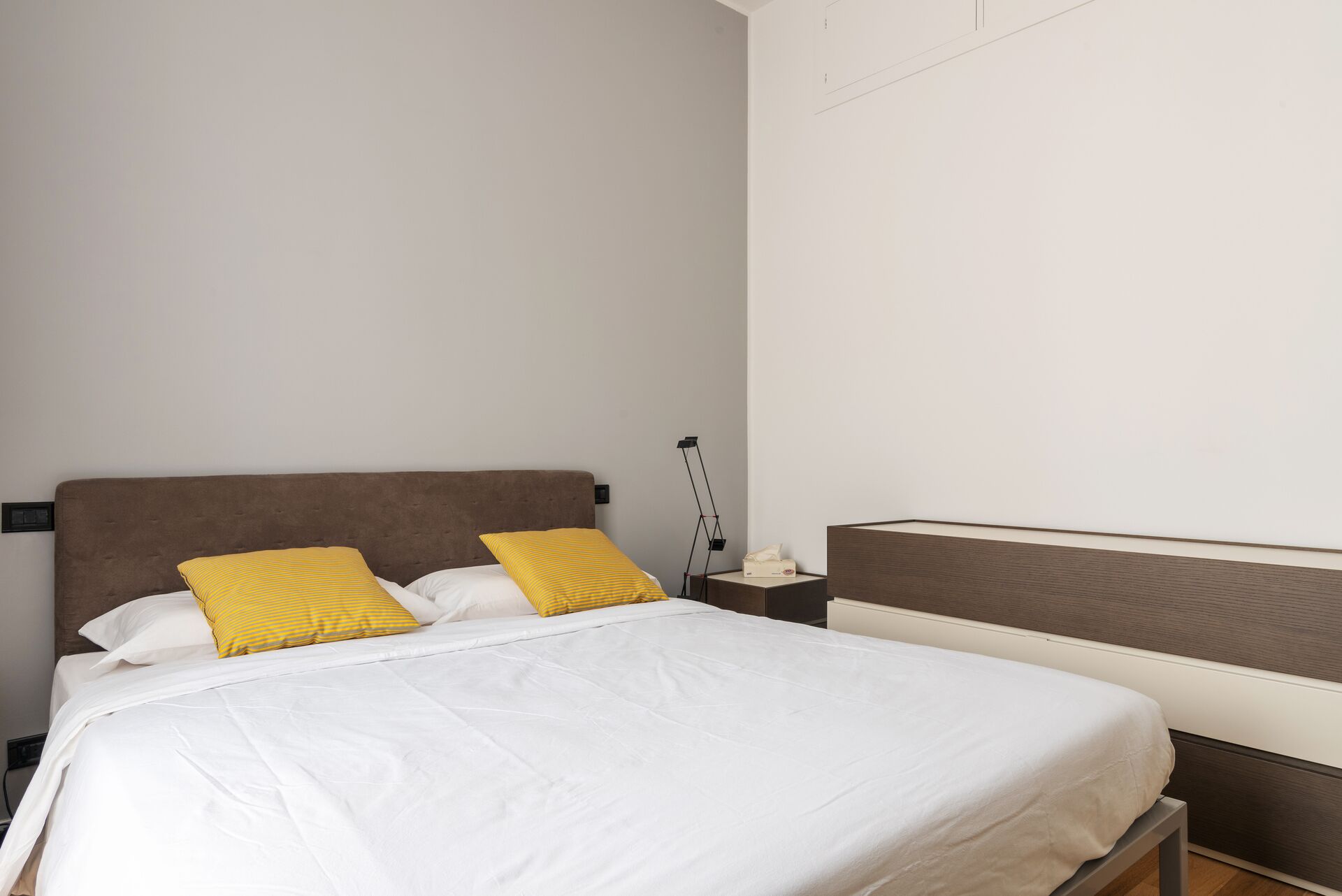 Cama bem arrumada com travesseiros listrados em branco e amarelo, cabeceira marrom e mesa de cabeceira com abajur e caixa de lenços.