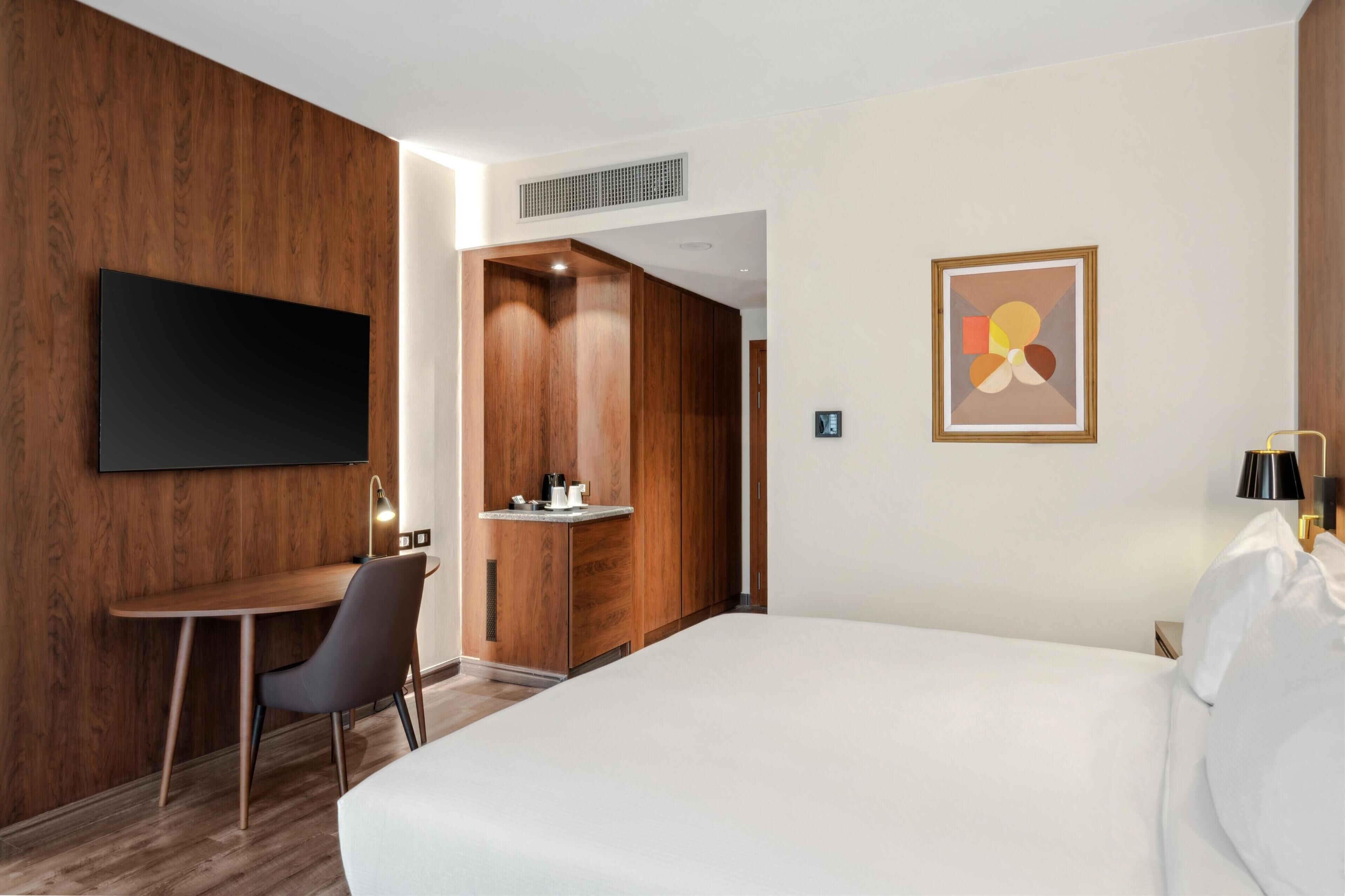 Quarto de hotel com cama, escrivaninha, cadeira, televisão, guarda-roupa e um quadro na parede.