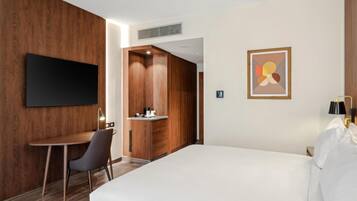 Quarto de hotel com cama, escrivaninha, cadeira, televisão, guarda-roupa e um quadro na parede.